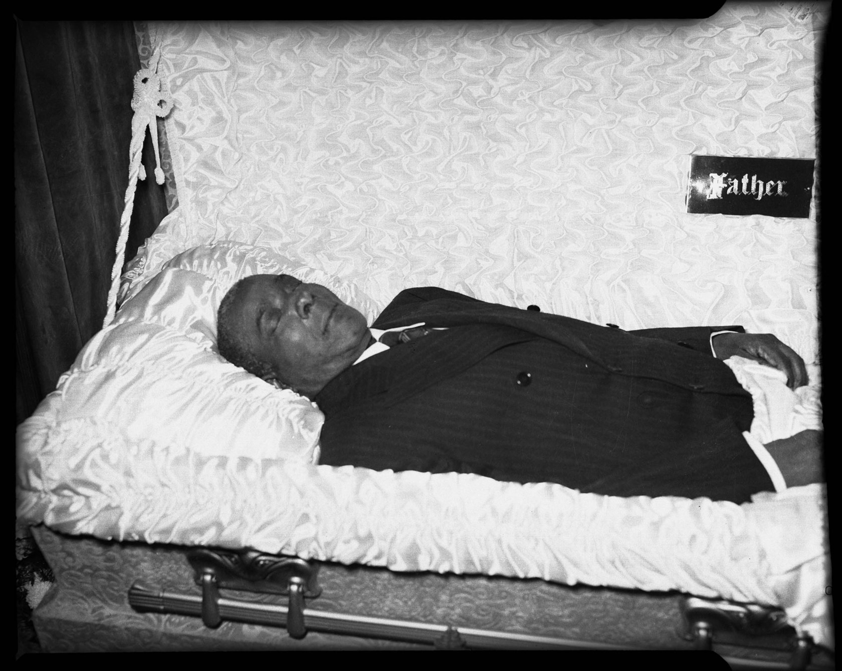 Deceased man wearing dark striped suit, lying in light colored floral ...